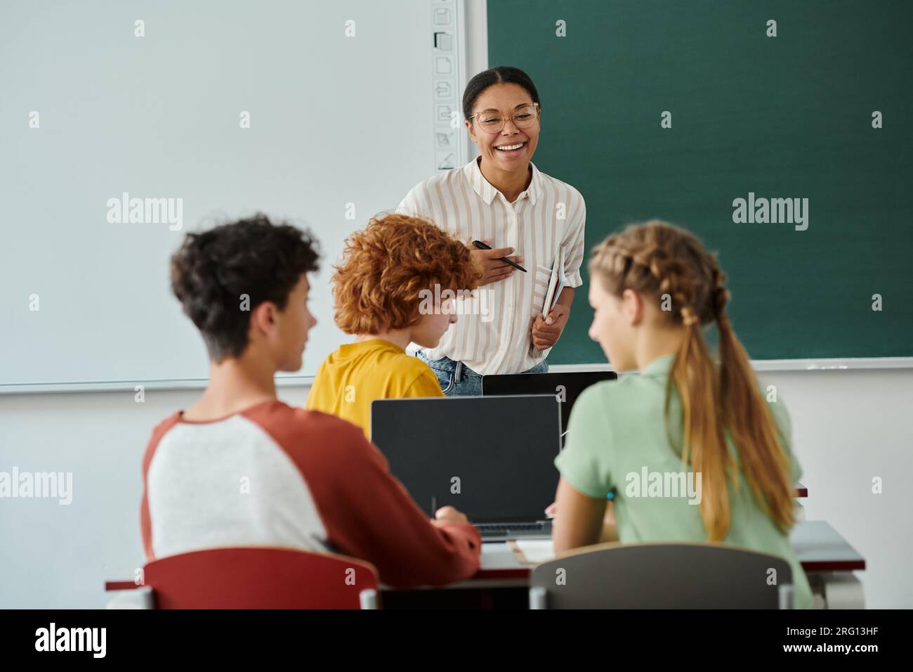African american teacher holding notebook near pupils with devices ...