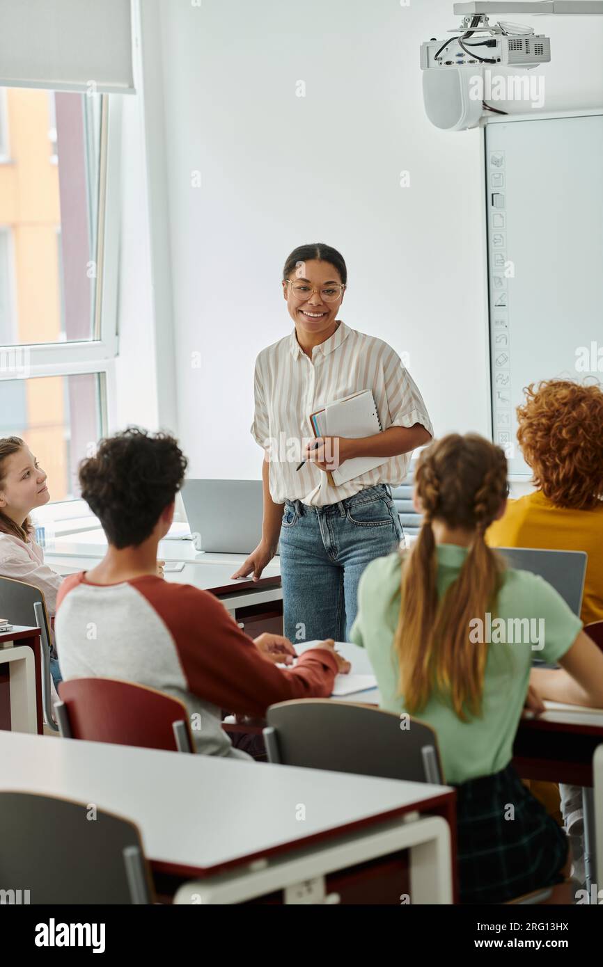 Young smiling african american teacher with notebook talking to pupils ...