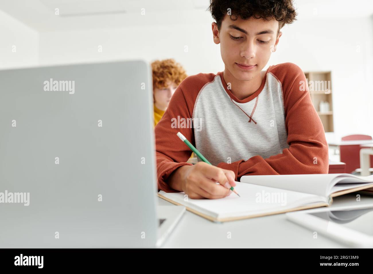 Teenage schoolboy writing on notebook near laptop and blurred classmate ...