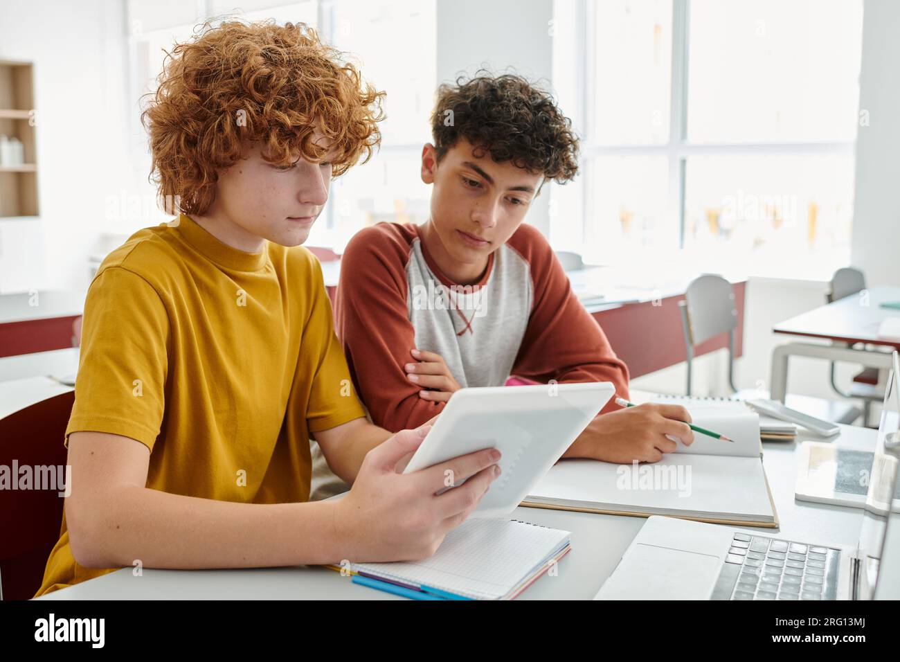 Teenage schoolboys using digital tablet together near notebooks on desk ...