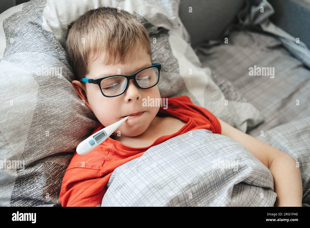 A boy with glasses lies on a bed with a thermometer in his mouth ...