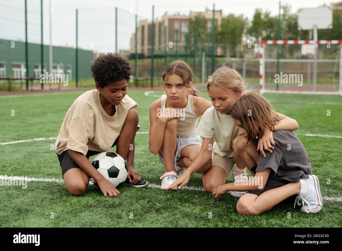 Four schoolchildren in activewear standing on knees on green football ...