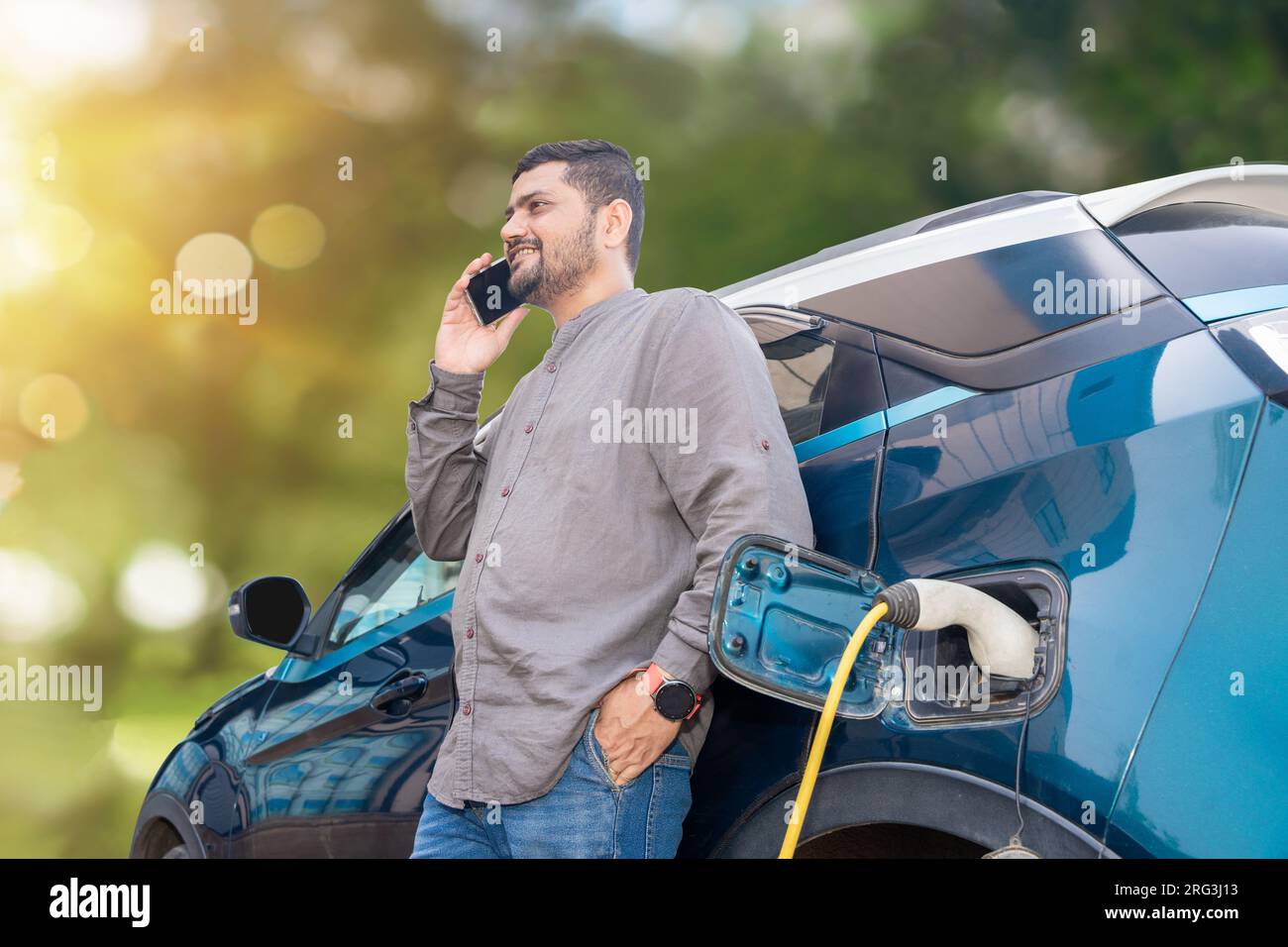 Happy Indian man talking on phone while his modern electric car getting ...