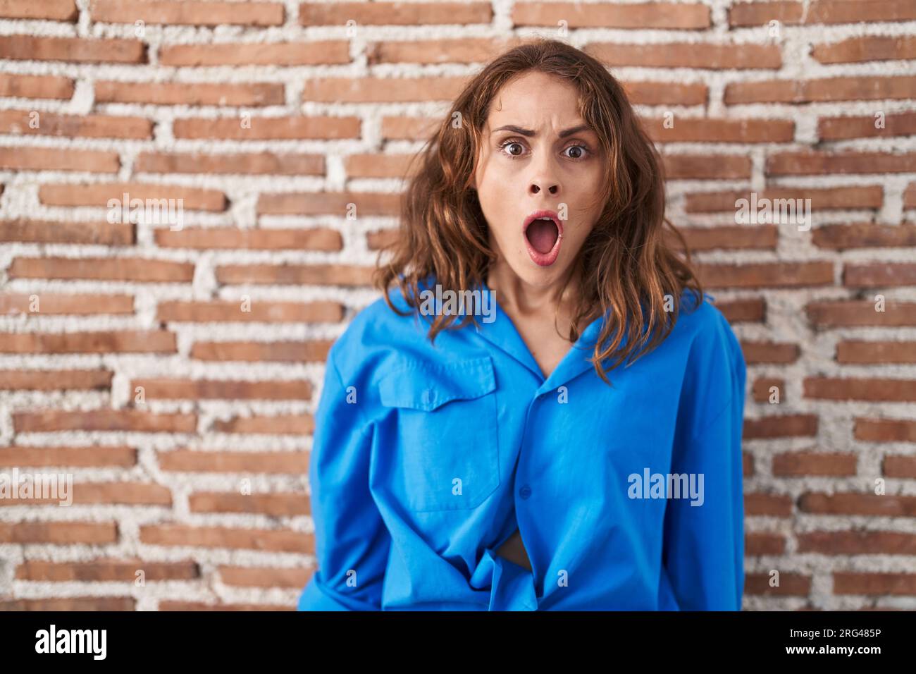 Beautiful brunette woman standing over bricks wall in shock face ...