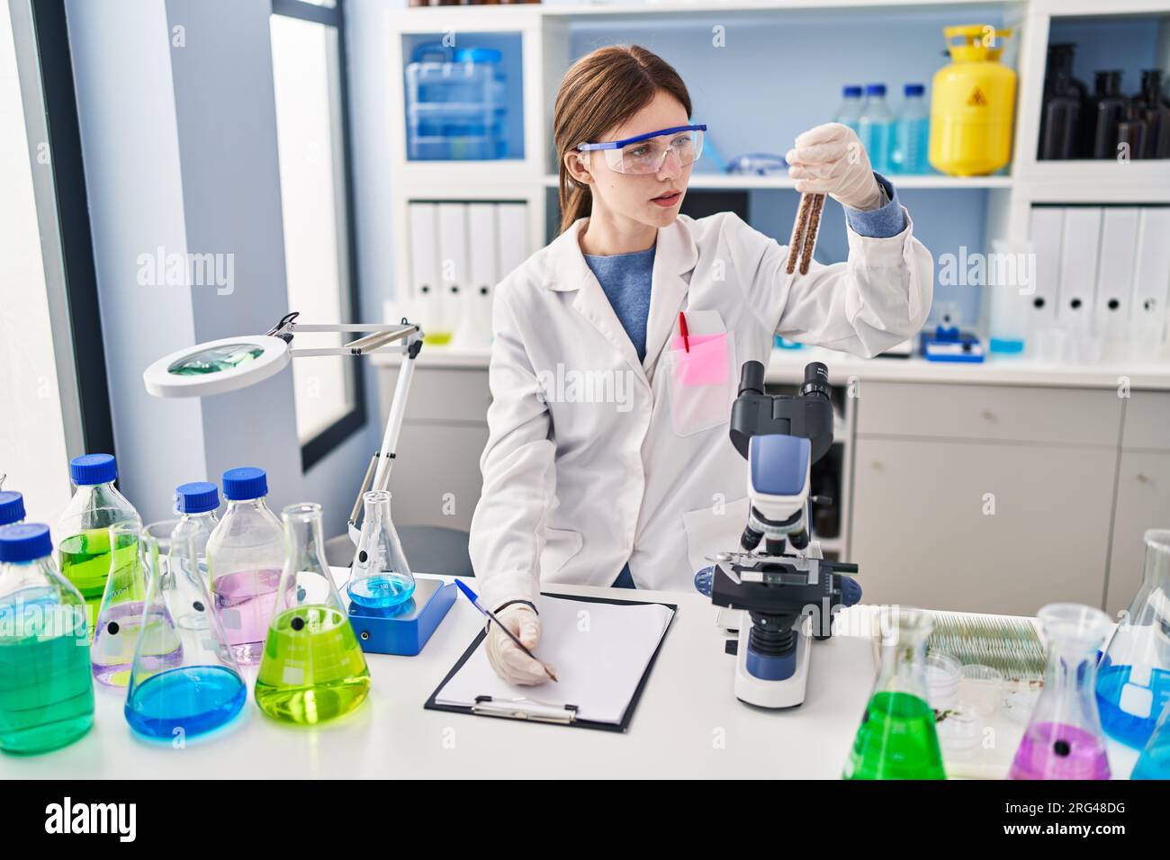 Young blonde woman scientist analysing test tube writing on clipboard ...