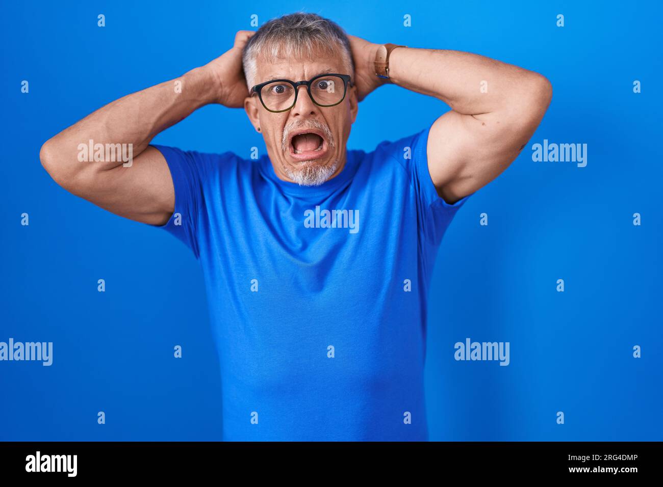 Hispanic man with grey hair standing over blue background crazy and ...