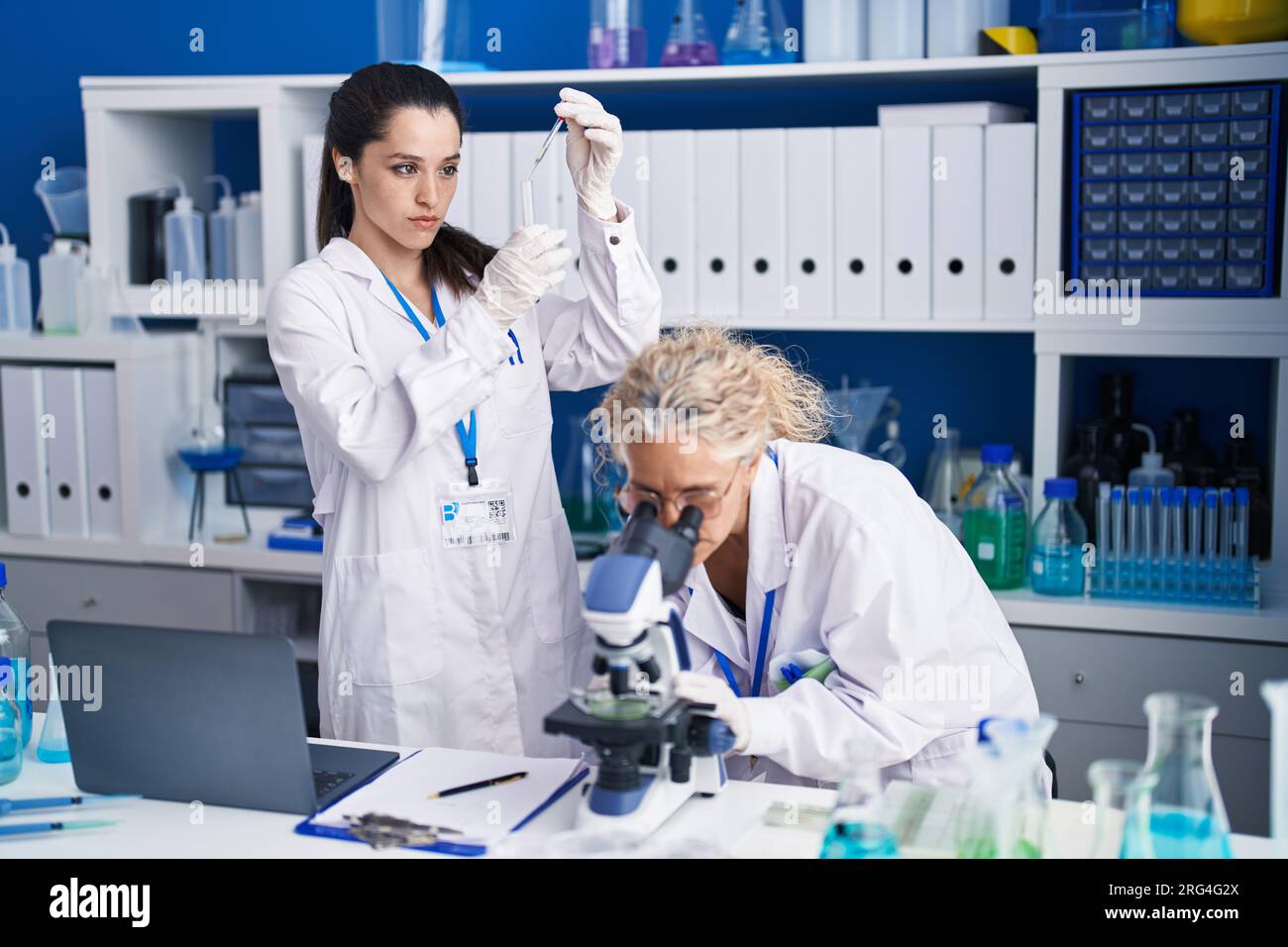 Two women scientists using microscope measuring liquid at laboratory ...