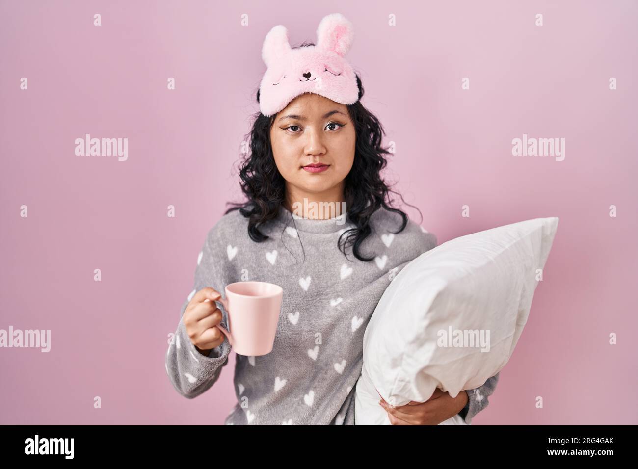 Young asian woman wearing sleep mask and pajama hugging pillow drinking ...