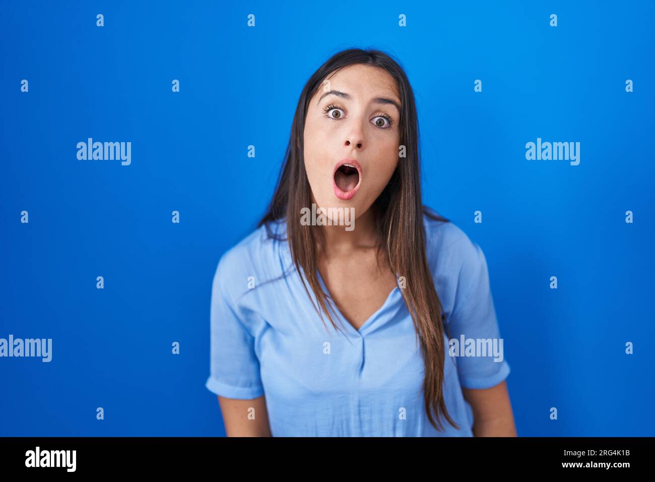 Young brunette woman standing over blue background in shock face ...
