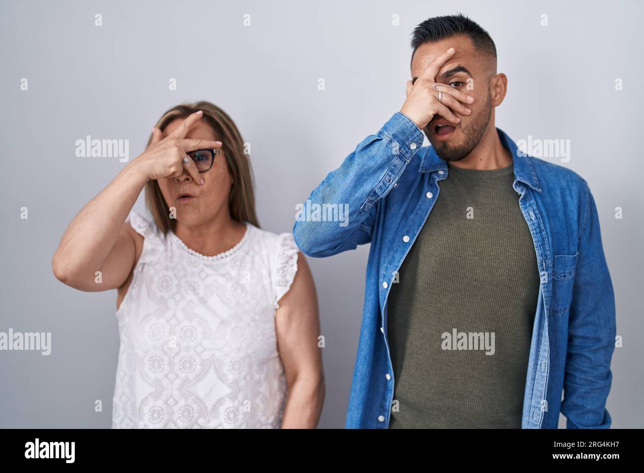 Hispanic mother and son standing together peeking in shock covering face and eyes with hand ...