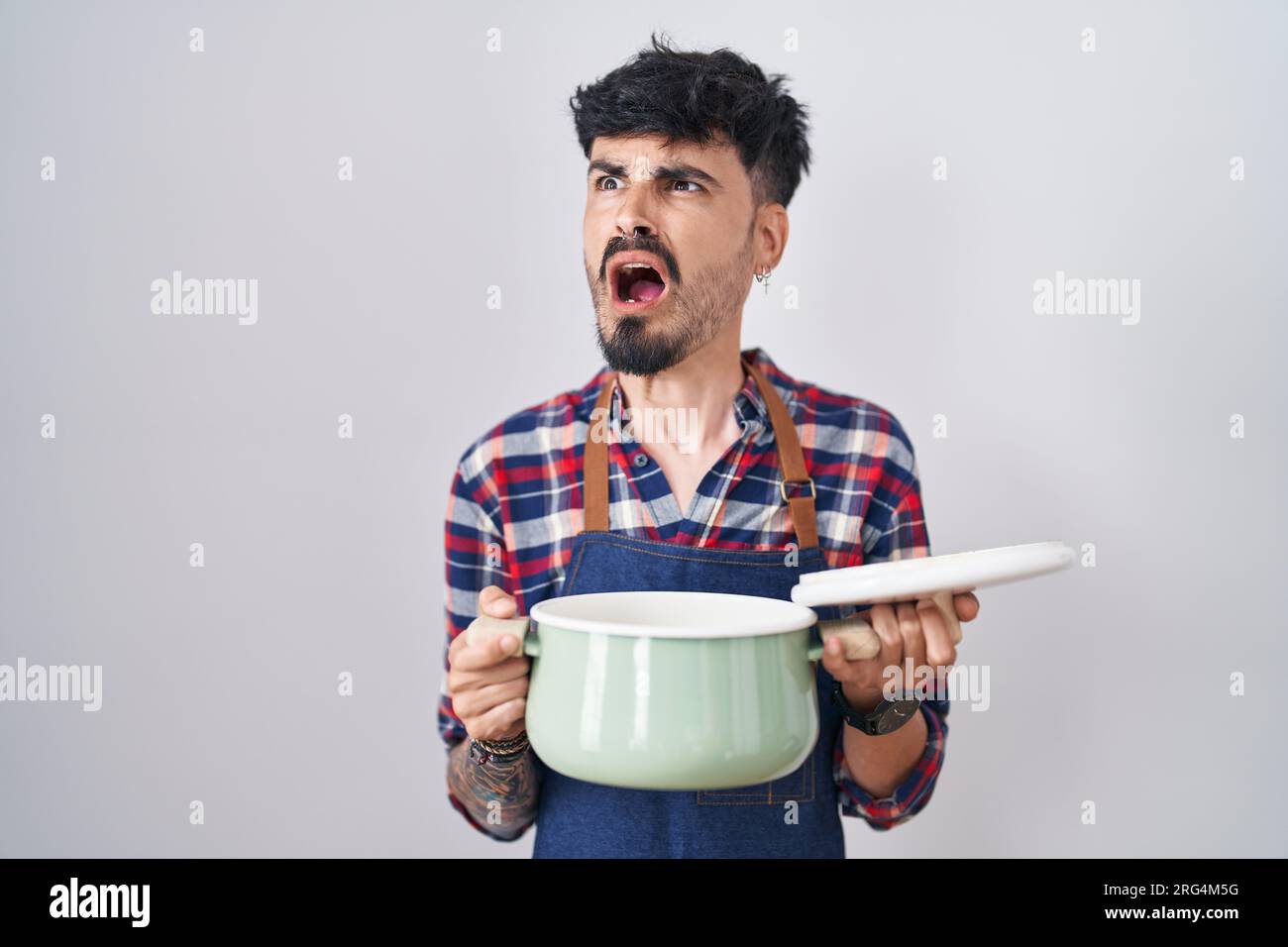 Young hispanic man with beard wearing apron holding cooking pot angry ...