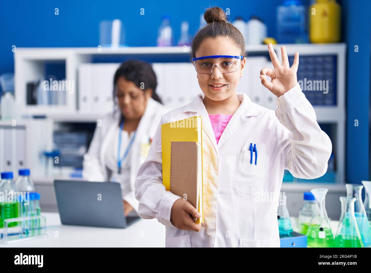 Mother and young daughter working at scientist laboratory doing ok sign with fingers, smiling ...