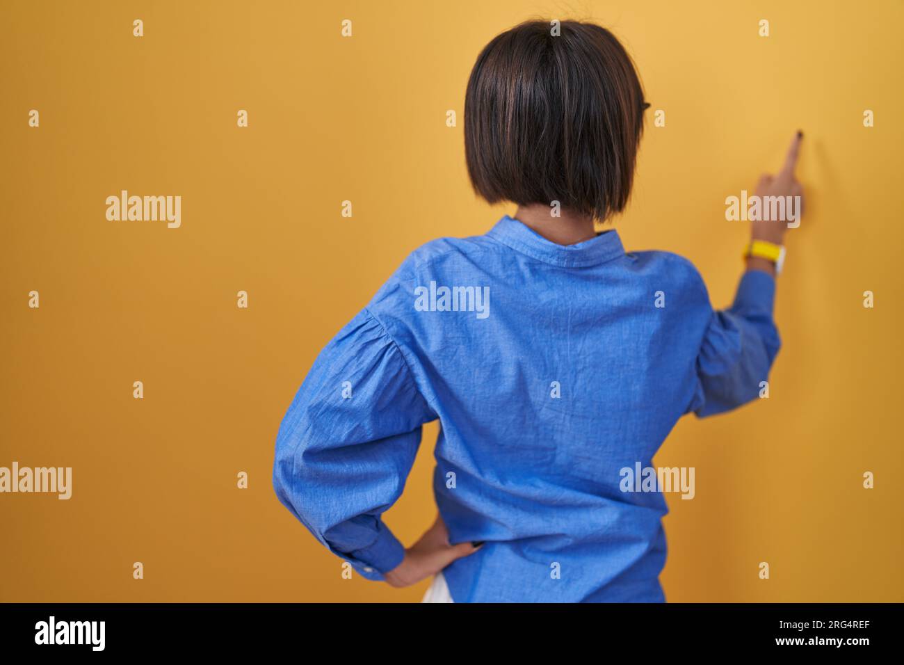 Young girl standing over yellow background posing backwards pointing ...