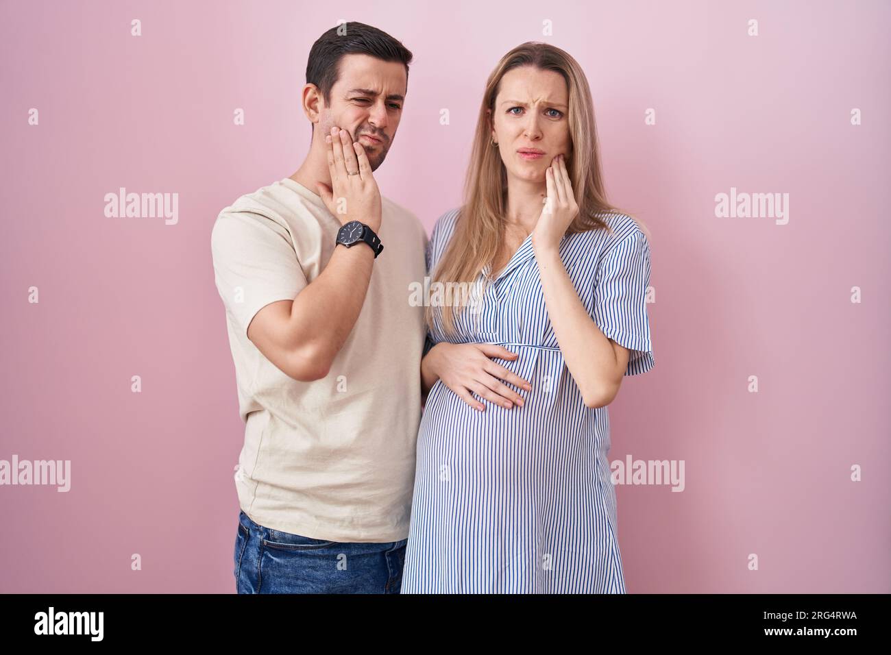 Young couple expecting a baby standing over pink background touching ...