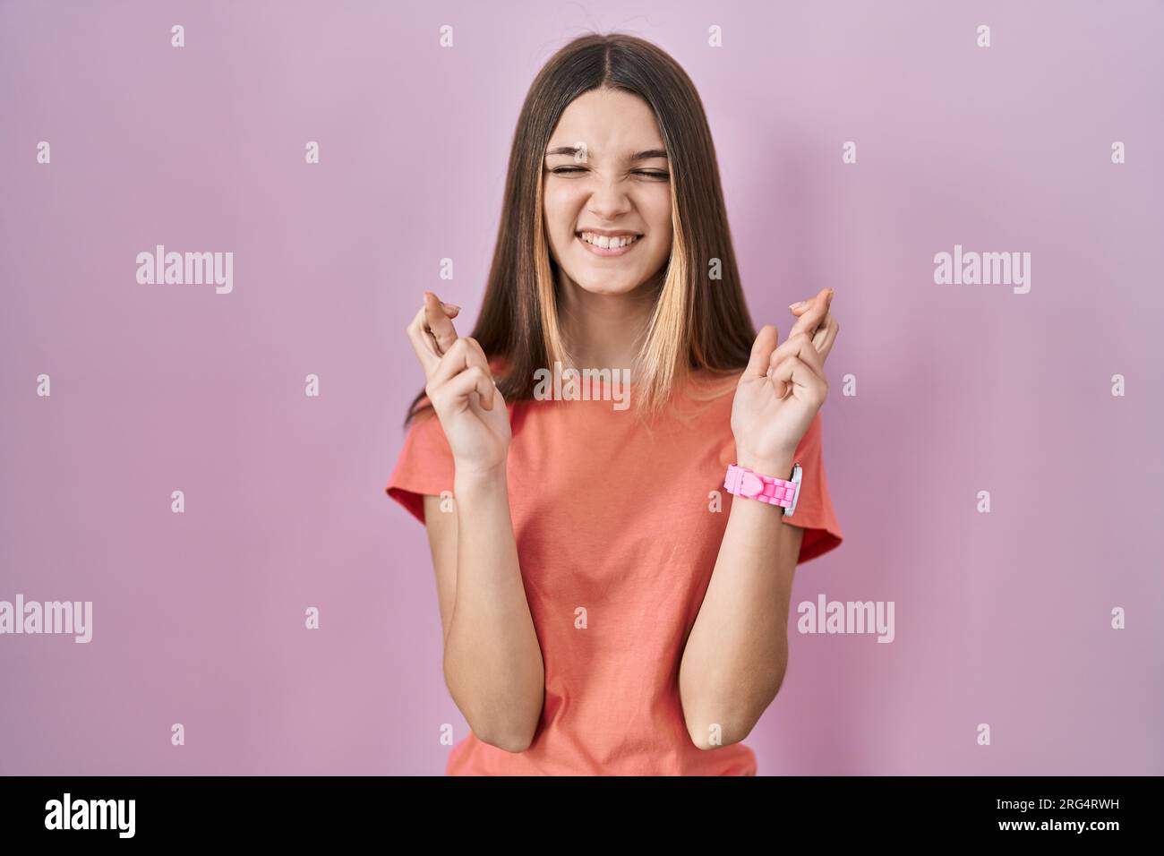Teenager girl standing over pink background gesturing finger crossed ...