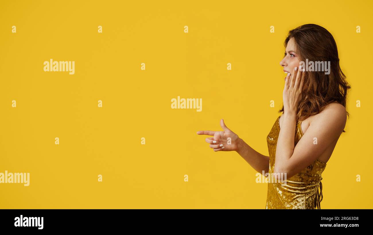 Shocked young woman pointing finger at empty space and touching cheek ...