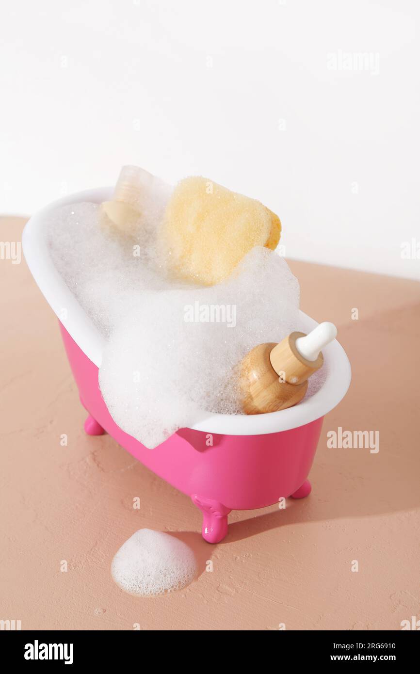 Small bathtub with foam, cosmetic products and sponge on color table ...