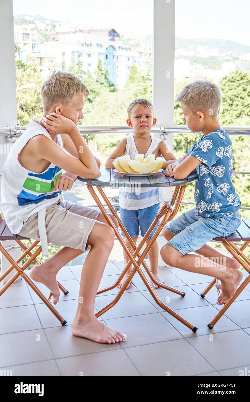 Preteen and junior and preschooler boys on hotel balcony with slices of fresh melon. Brothers ...