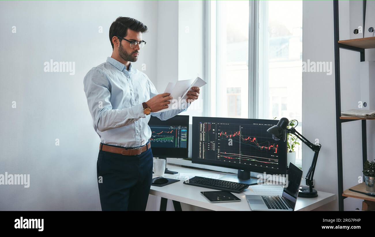 Analyzing data. Thoughtful trader in eyeglasses holding a financial report and analyzing trading ...