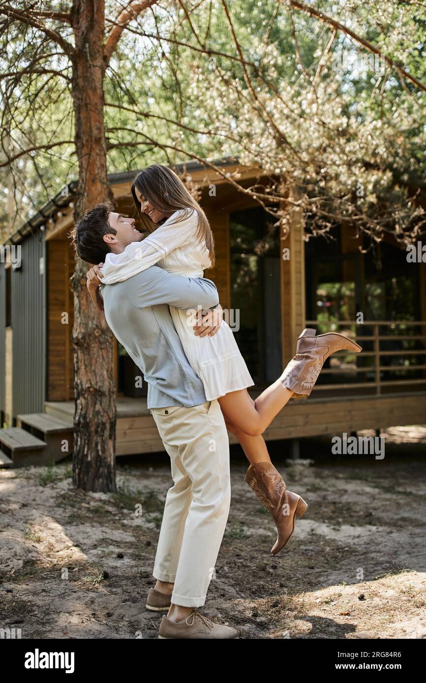 happy man lifting and hugging woman in vacation house, forest, look at ...