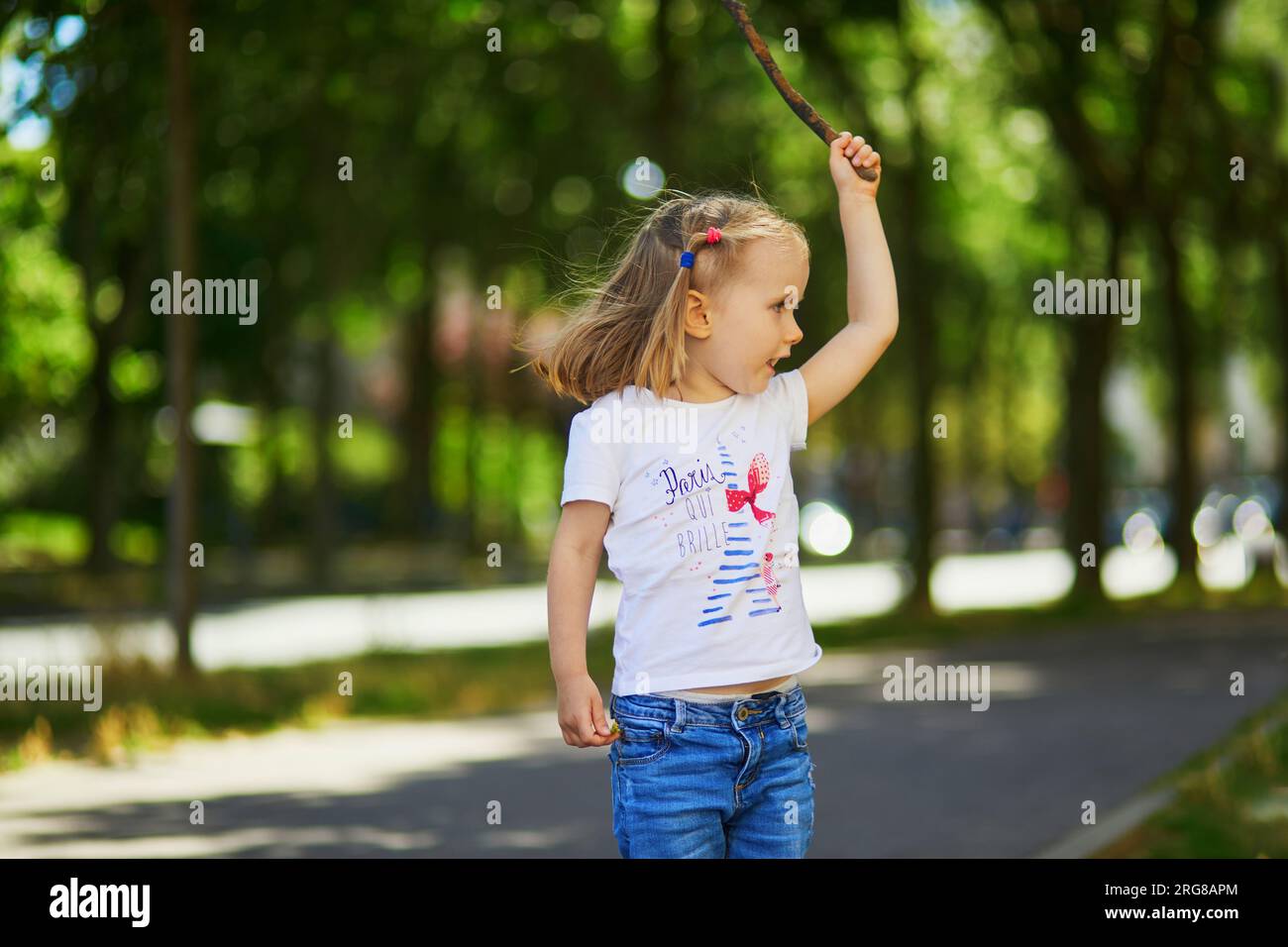 Adorable preschooler girl playing with a stick on a sunny street ...