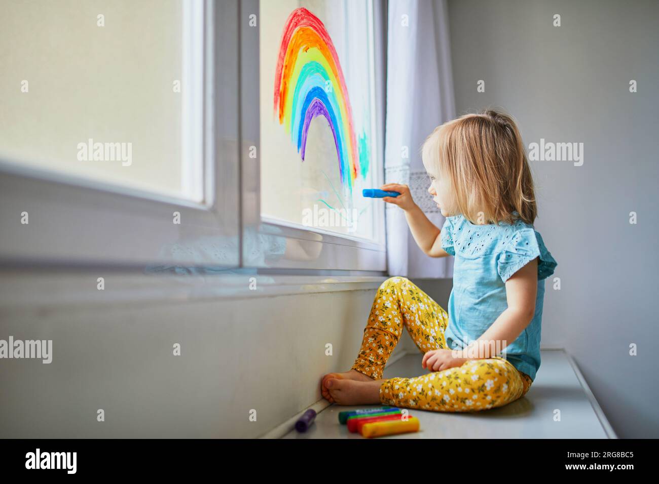 Adorable toddler girl drawing rainbow on window glass as sign of hope ...