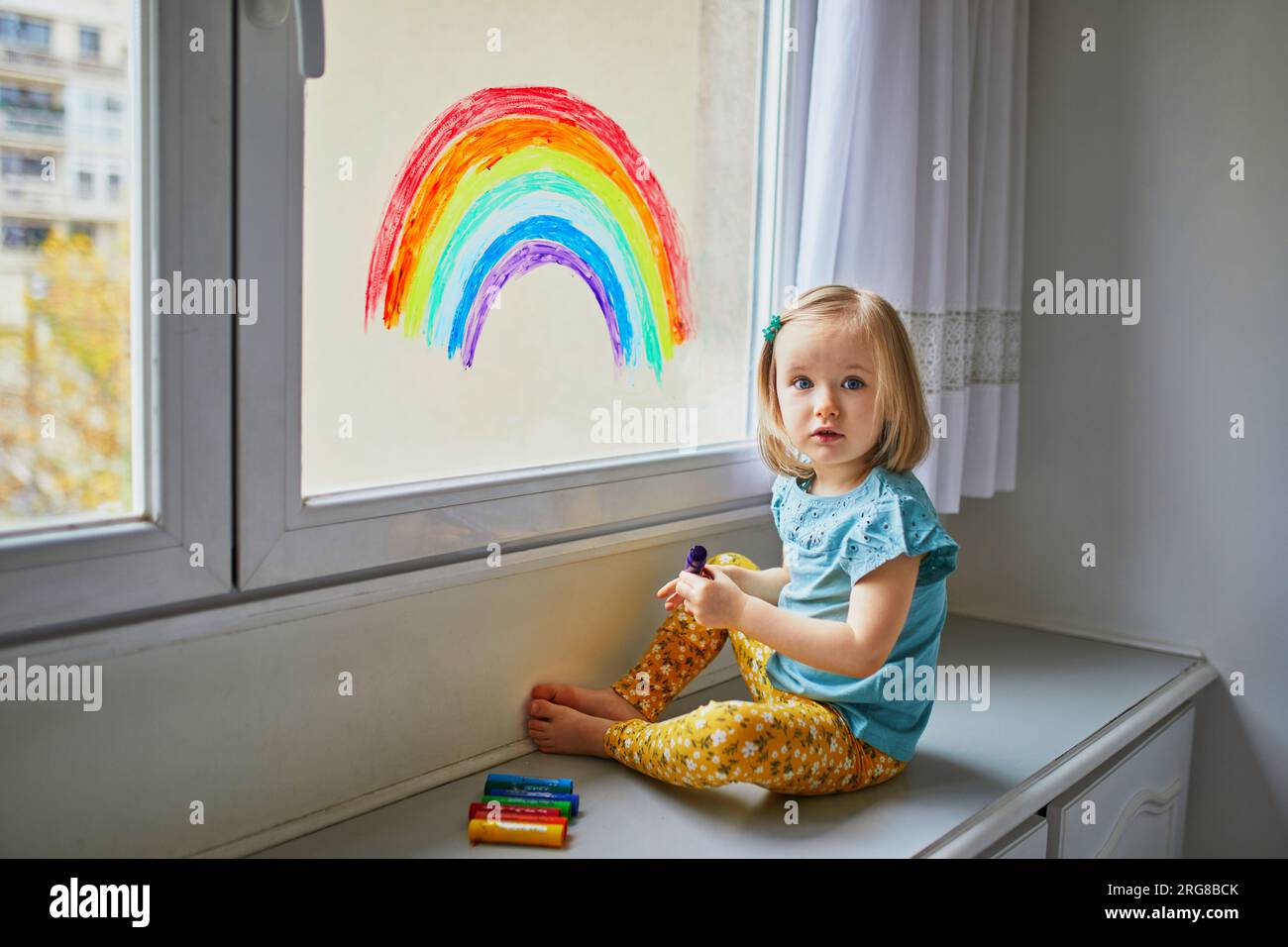 Adorable toddler girl drawing rainbow on window glass as sign of hope ...