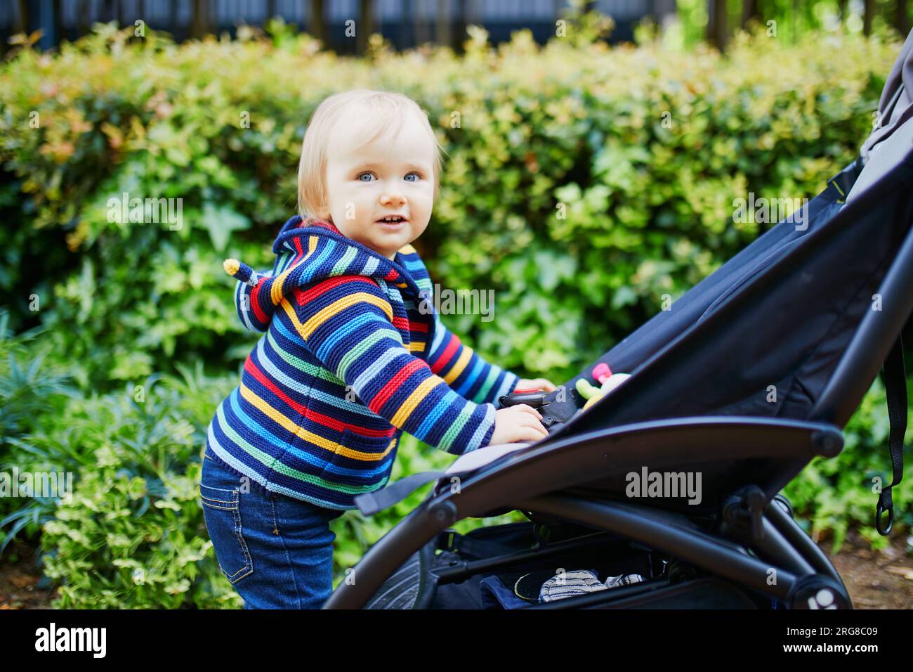 Adorable happy toddler girl standing next to her pushchair in park ...