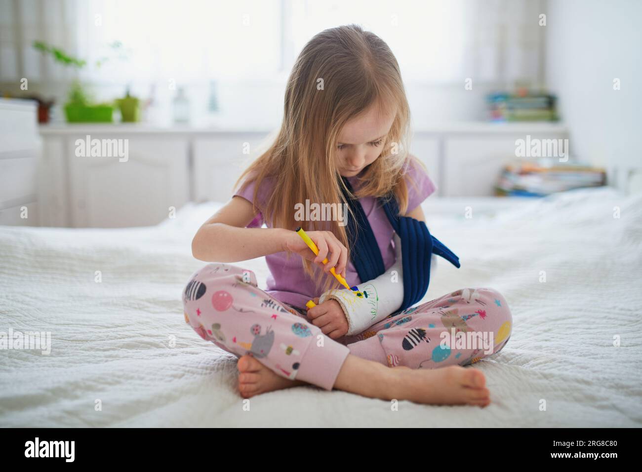 Adorable preschooler girl with a broken arm at home on the bed draws ...