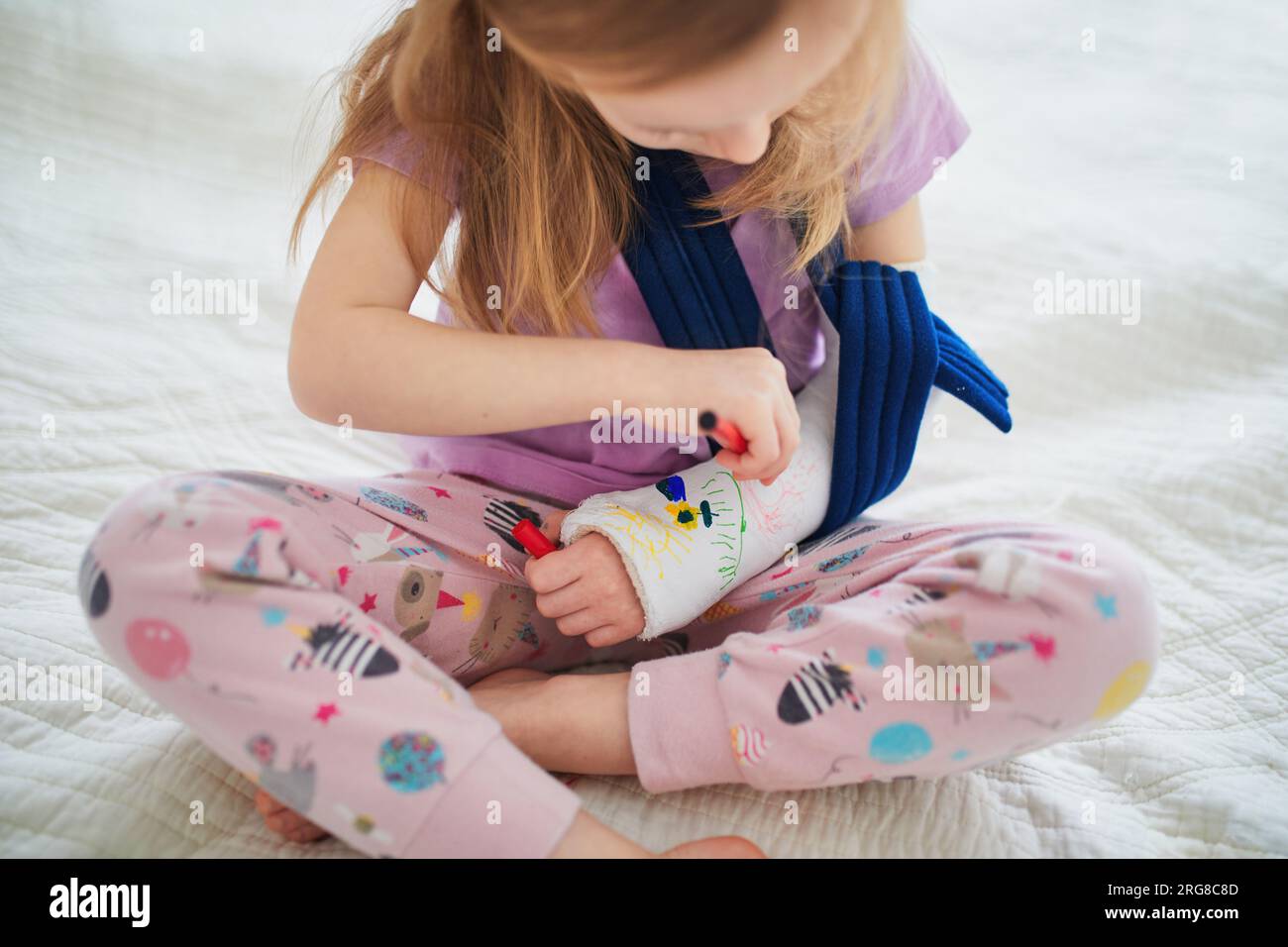 Adorable preschooler girl with a broken arm at home on the bed draws ...