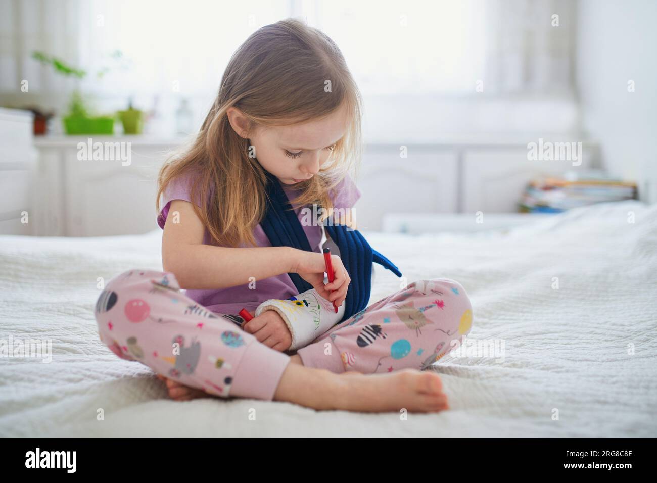 Adorable preschooler girl with a broken arm at home on the bed draws ...