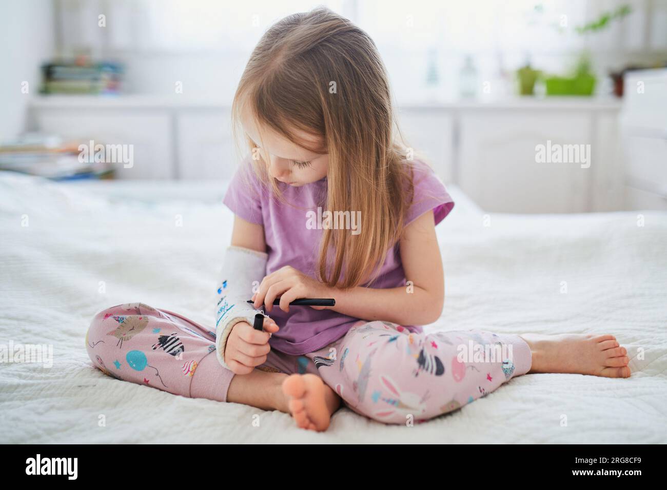 Adorable preschooler girl with a broken arm at home on the bed draws ...
