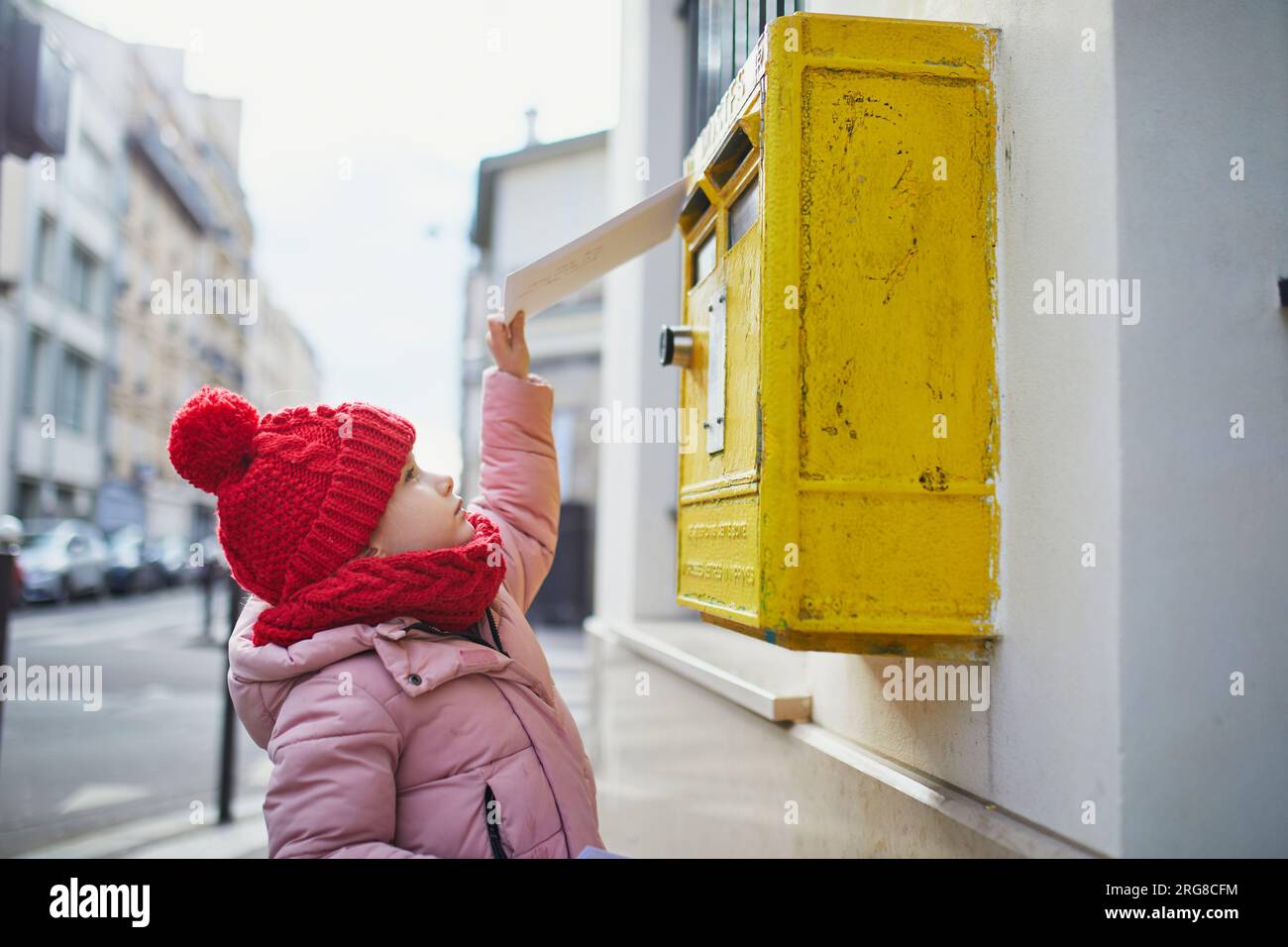 Adorable preschooler girl putting letter in yellow post box on a street ...