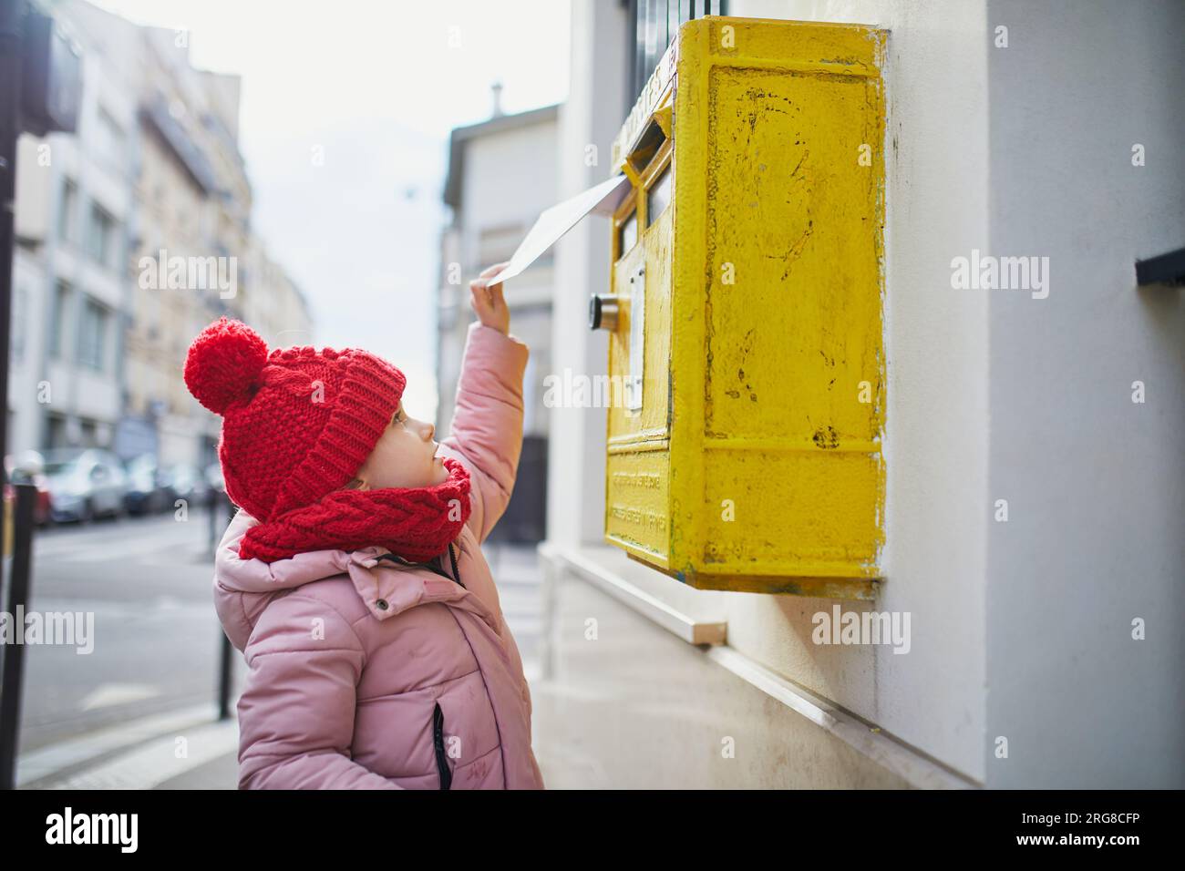 Adorable preschooler girl putting letter in yellow post box on a street ...