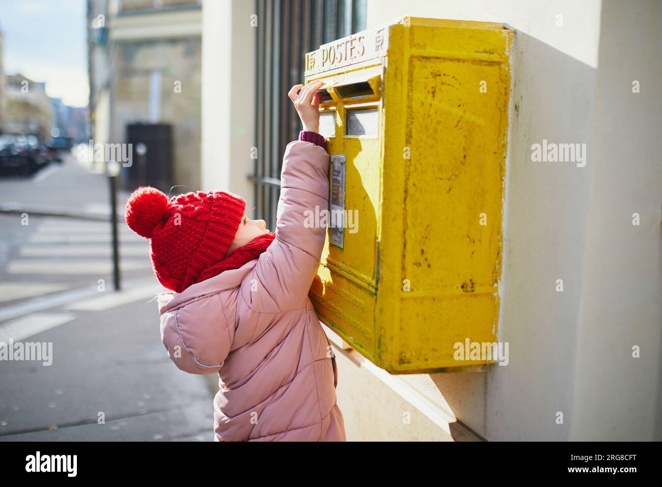 Adorable preschooler girl putting letter in yellow post box on a street ...
