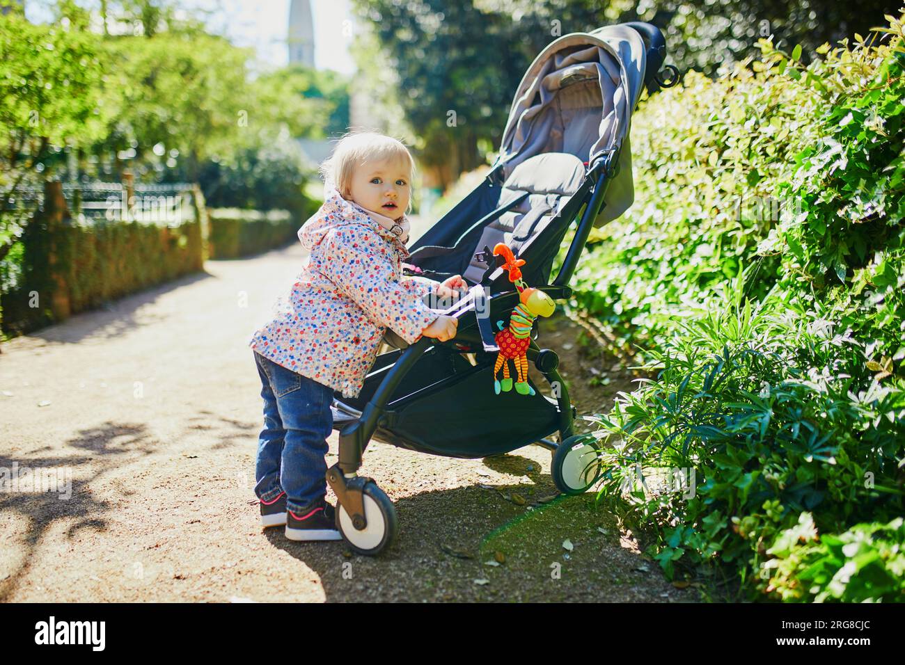 Adorable happy toddler girl standing next to her pushchair in park ...