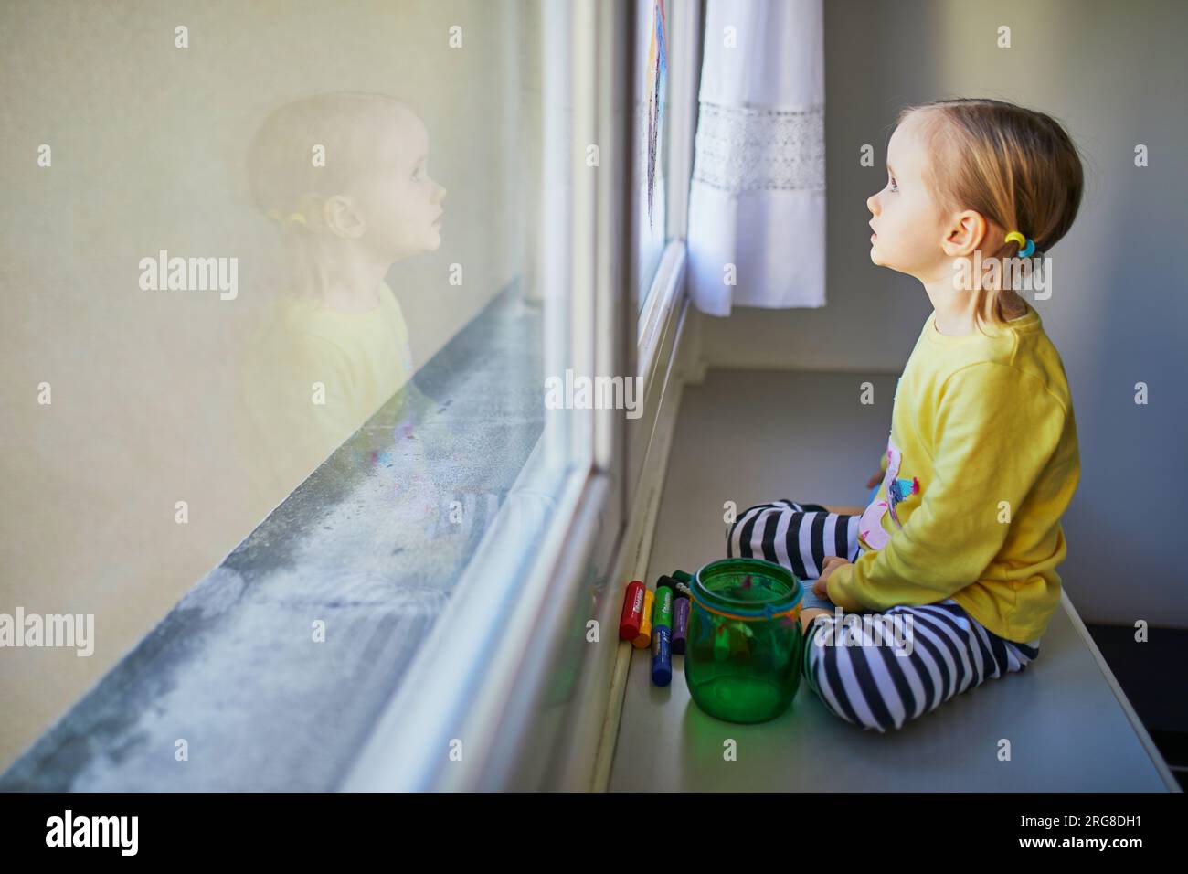 Adorable toddler girl sitting on window sill and looking out of the ...