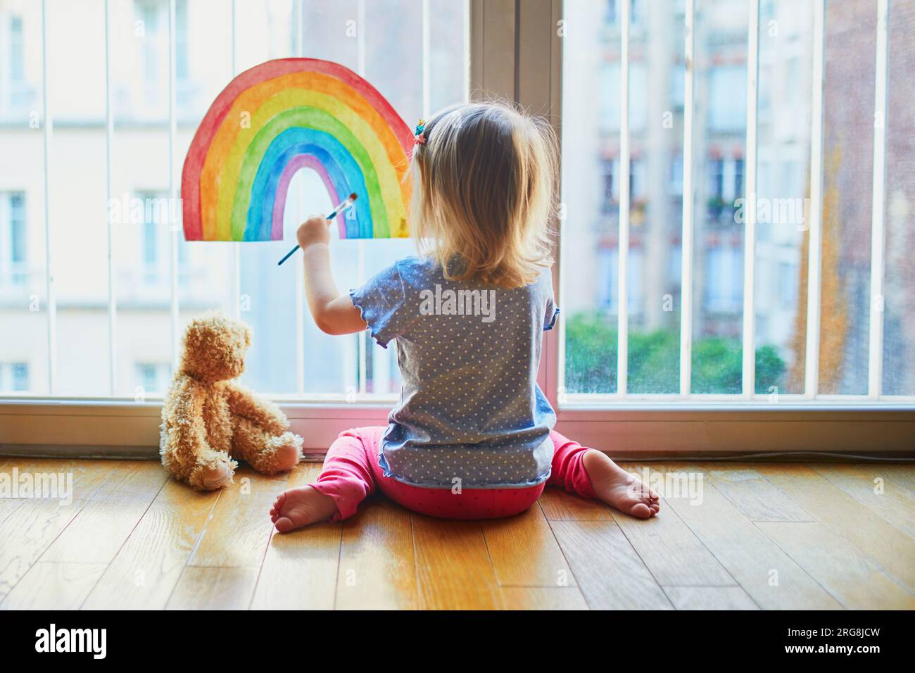 Adorable toddler girl drawing rainbow on window glass as sign of hope ...