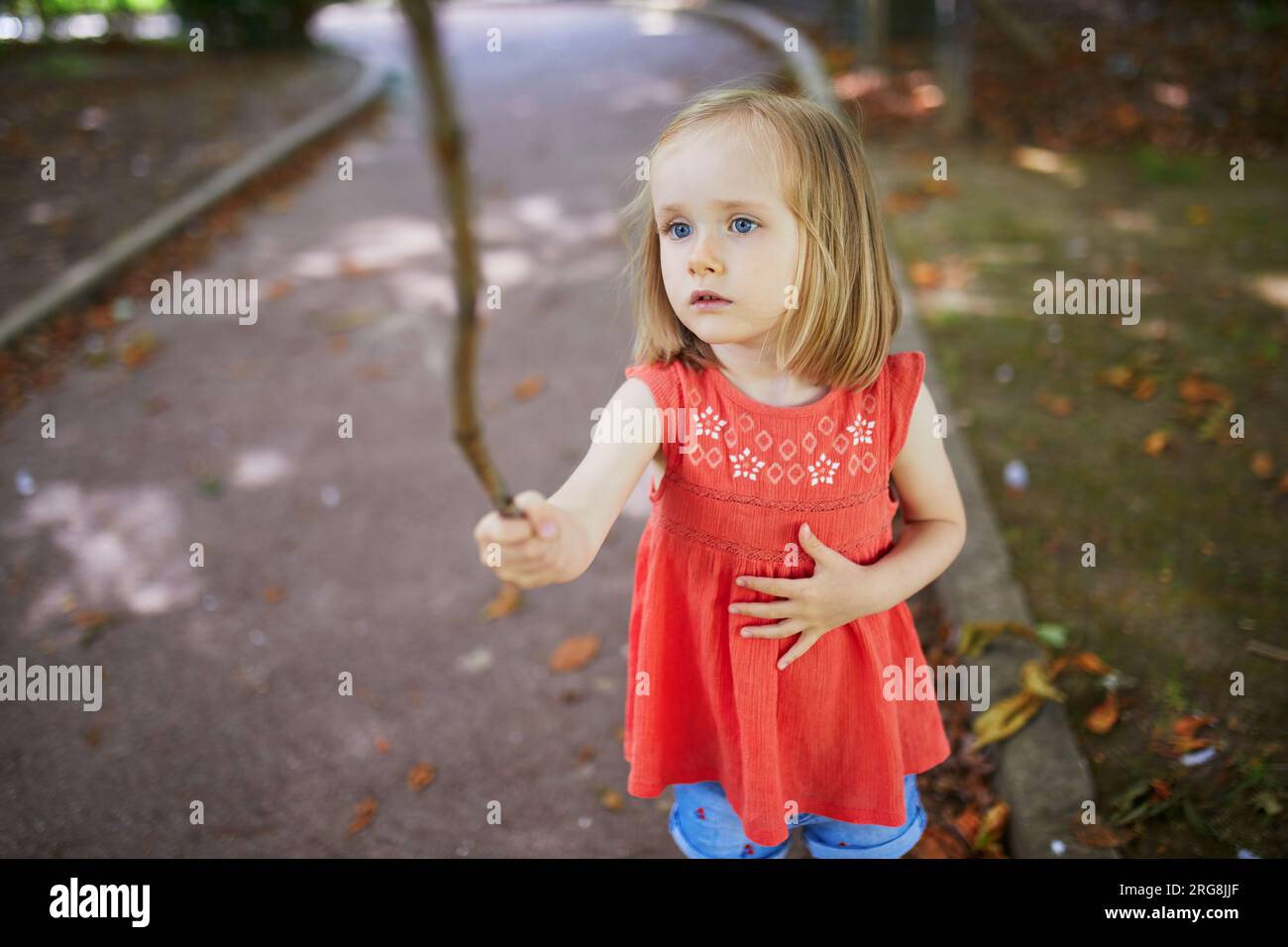 Adorable preschooler girl playing with a stick on a sunny street ...