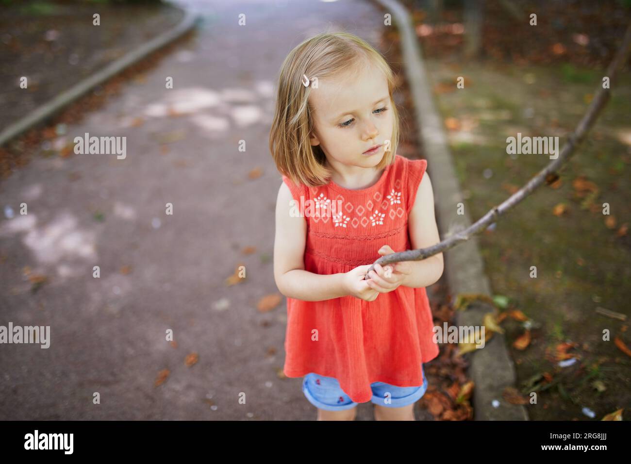 Adorable preschooler girl playing with a stick on a sunny street ...