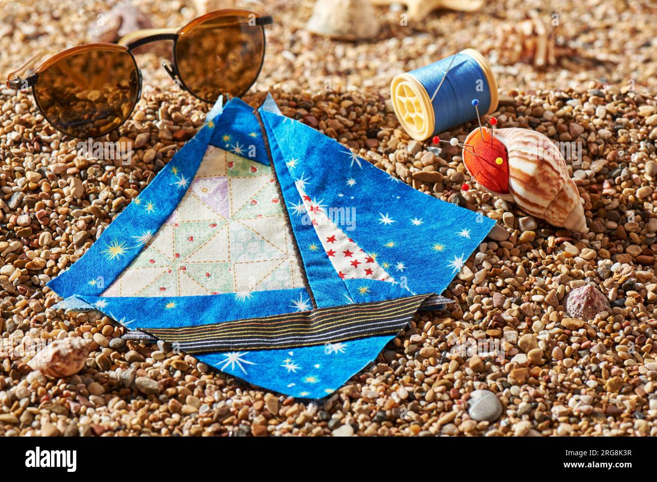 Patchwork block, spool of thread, pin cushion and sunglasses lie on sea stones of beach Stock ...