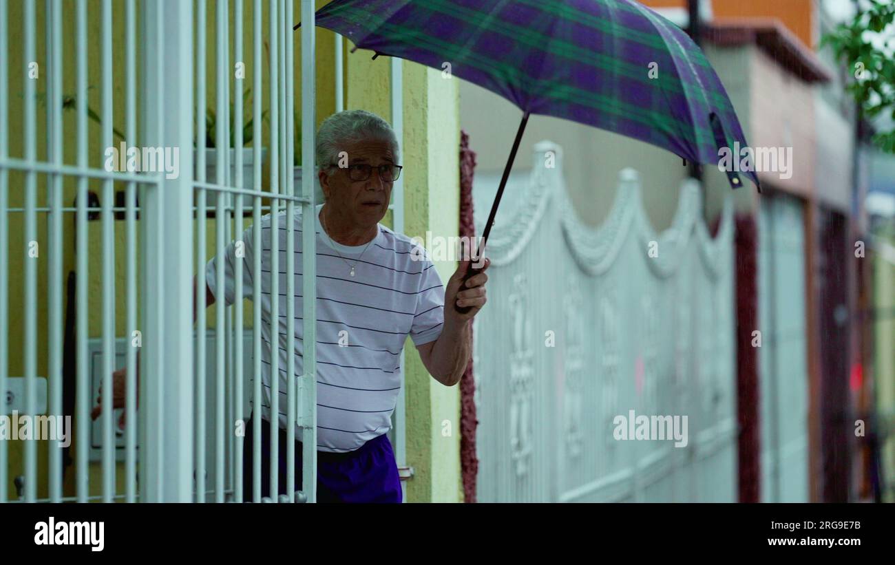 Older man leaving home while raining, opens umbrella and closes gate ...