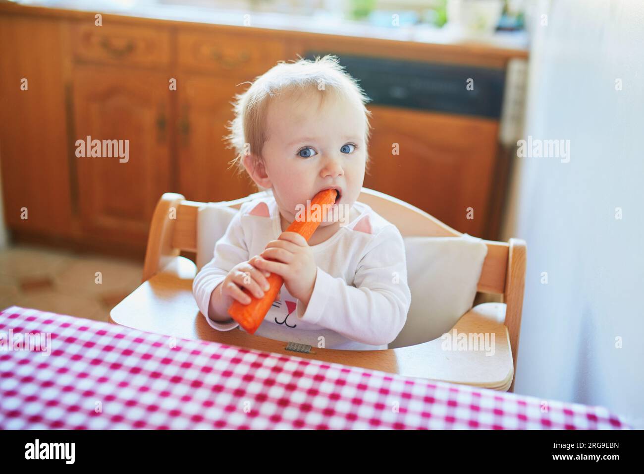 Cute baby girl eating carrot in the kitchen. Little kid tasting solids ...