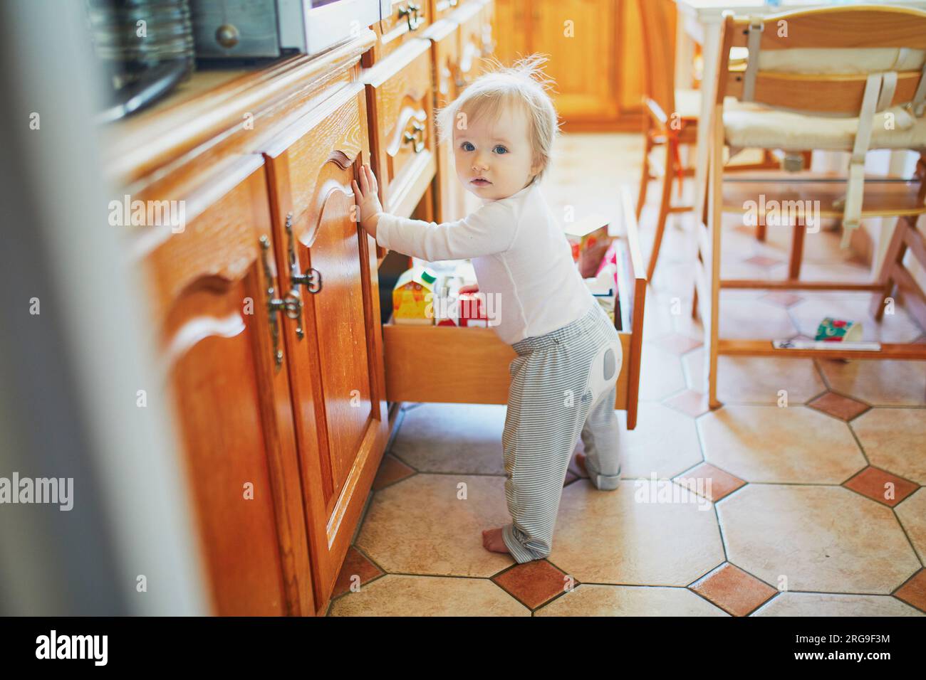 Adorable toddler girl at home, opening the drawer in the kitchen and ...