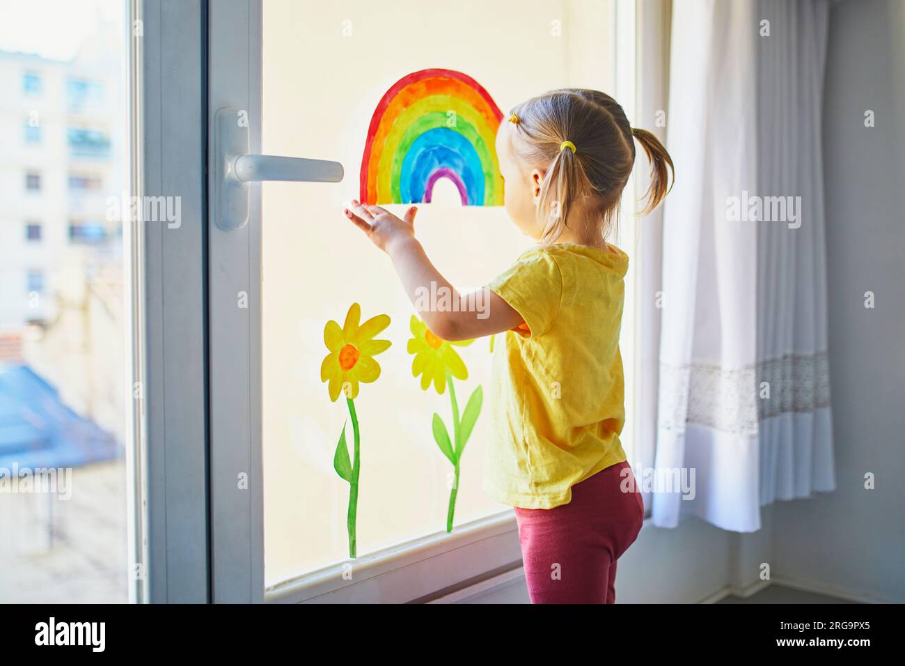 Adorable toddler girl attaching drawing of rainbow to window glass as ...