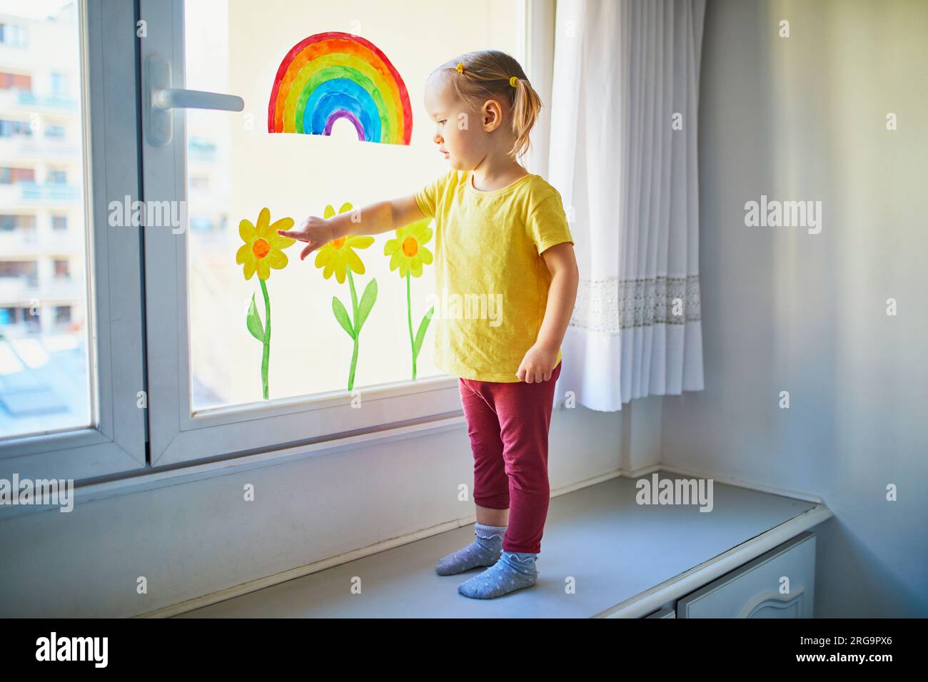Adorable toddler girl attaching drawing of rainbow to window glass as ...