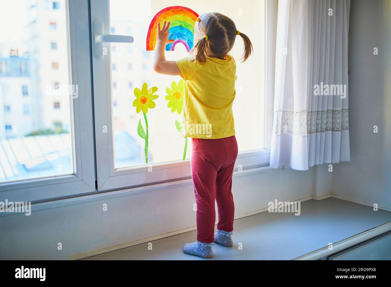 Adorable toddler girl attaching drawing of rainbow to window glass as ...
