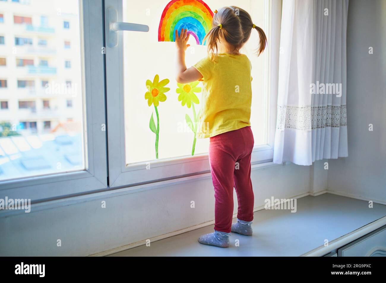 Adorable toddler girl attaching drawing of rainbow to window glass as ...