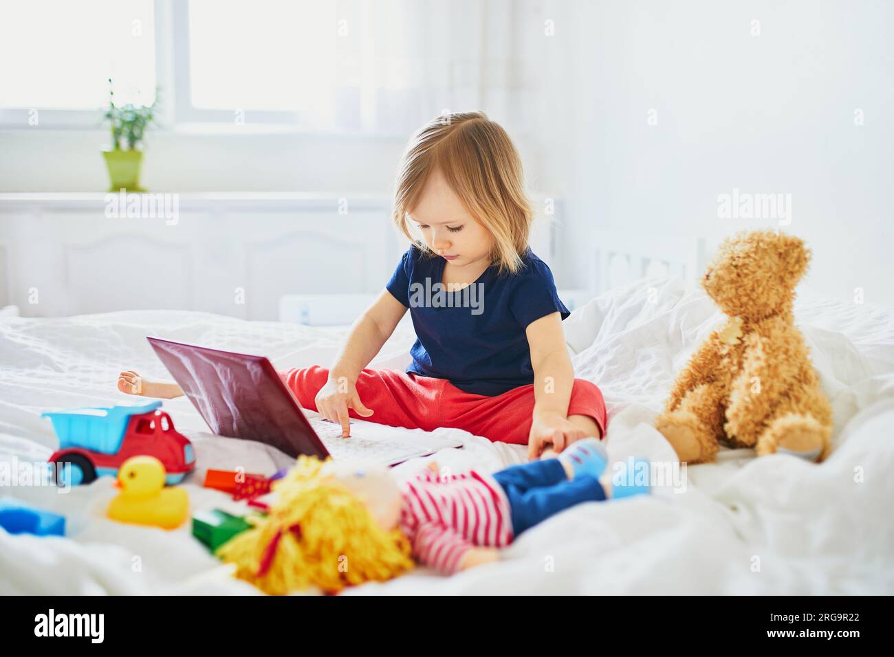 Toddler girl with laptop and toys in bed. Kid using gadget to ...