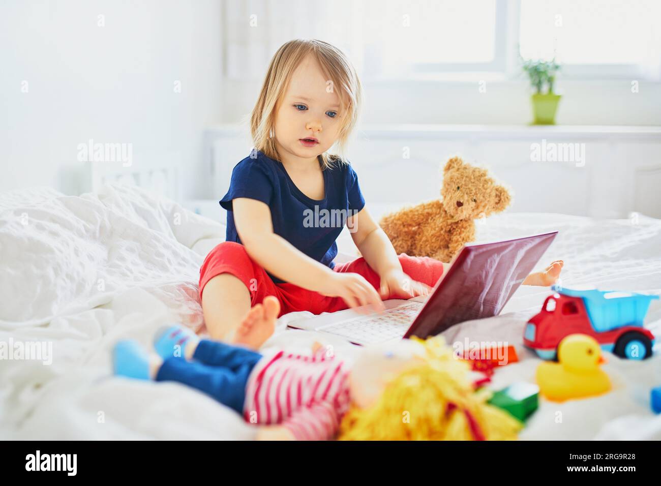 Toddler girl with laptop and toys in bed. Kid using gadget to ...